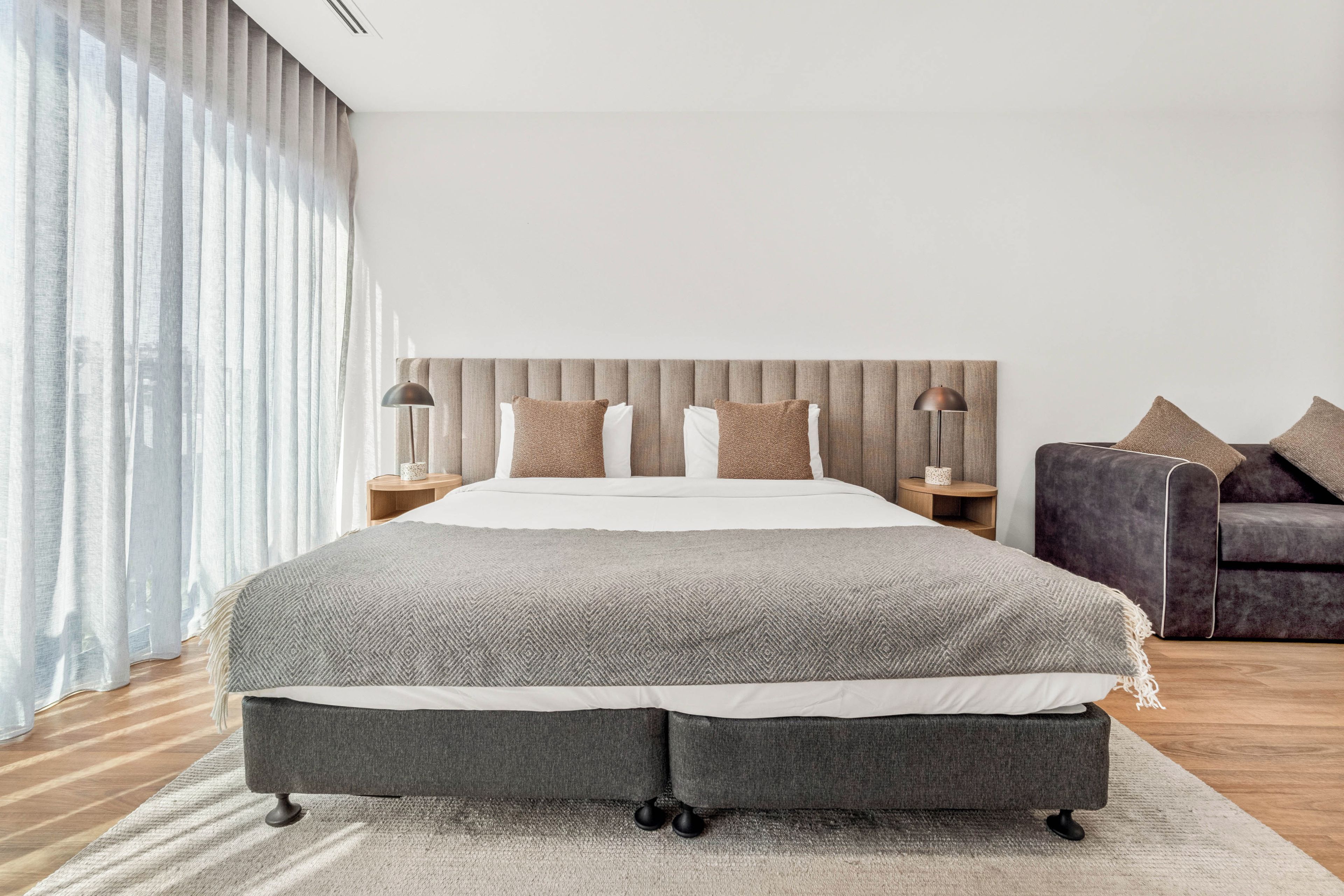 Quarto moderno com uma cama grande, um sofá estofado cinza e piso de madeira.