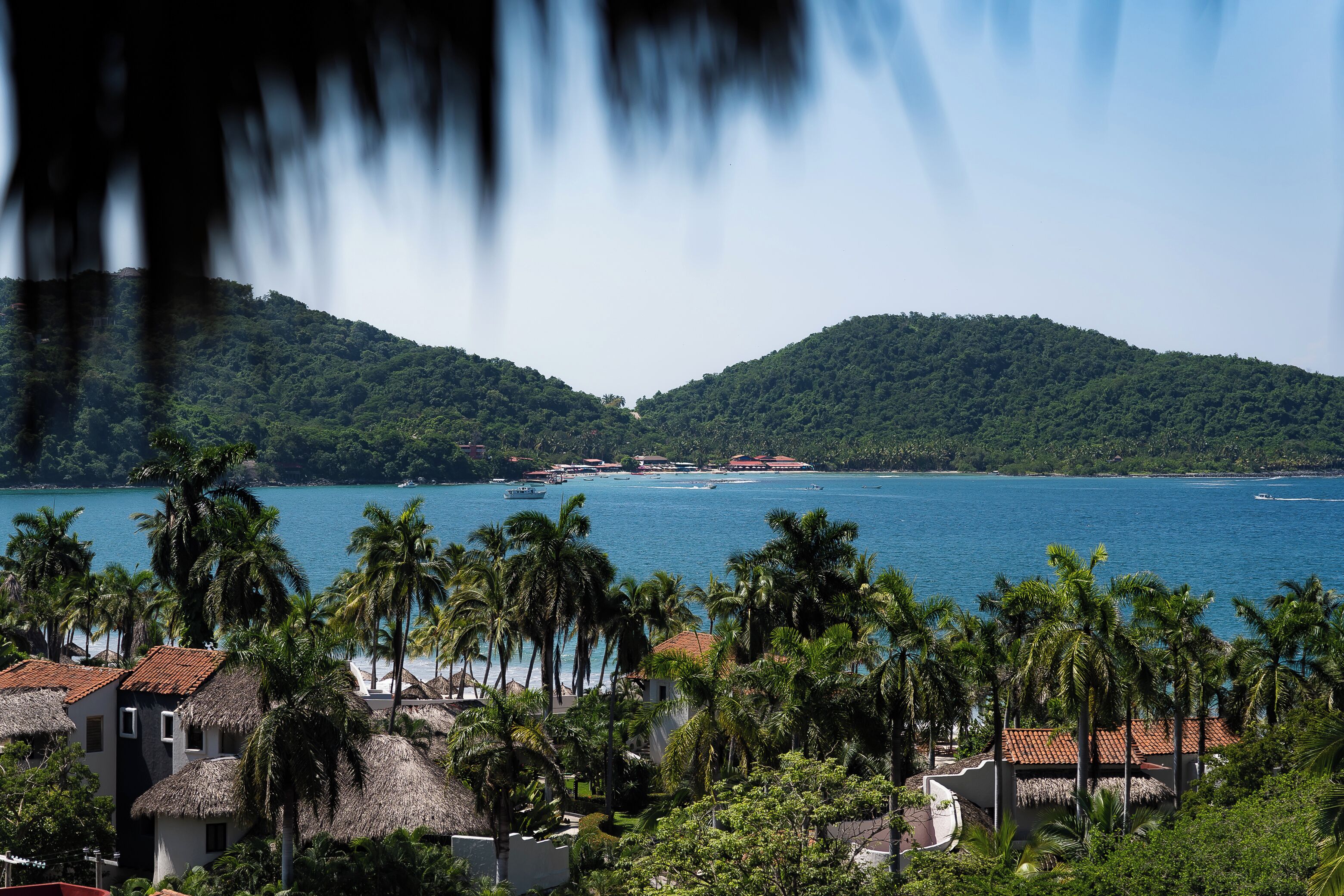 Uma vila costeira com casas de telhado de palha, palmeiras e vista para o mar e colinas.