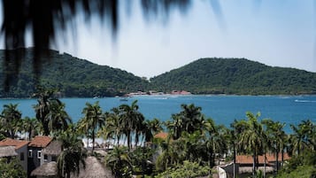 Uma vila costeira com casas de telhado de palha, palmeiras e vista para o mar e colinas.