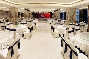 Uma sala de banquetes com mesas redondas e cadeiras, um palco com fundo vermelho e piso de mármore.