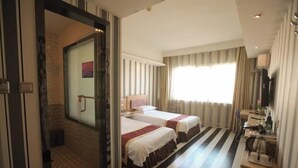 Quarto de hotel com duas camas, uma janela grande com cortinas, uma mesinha com chaleira e uma televisão na parede.