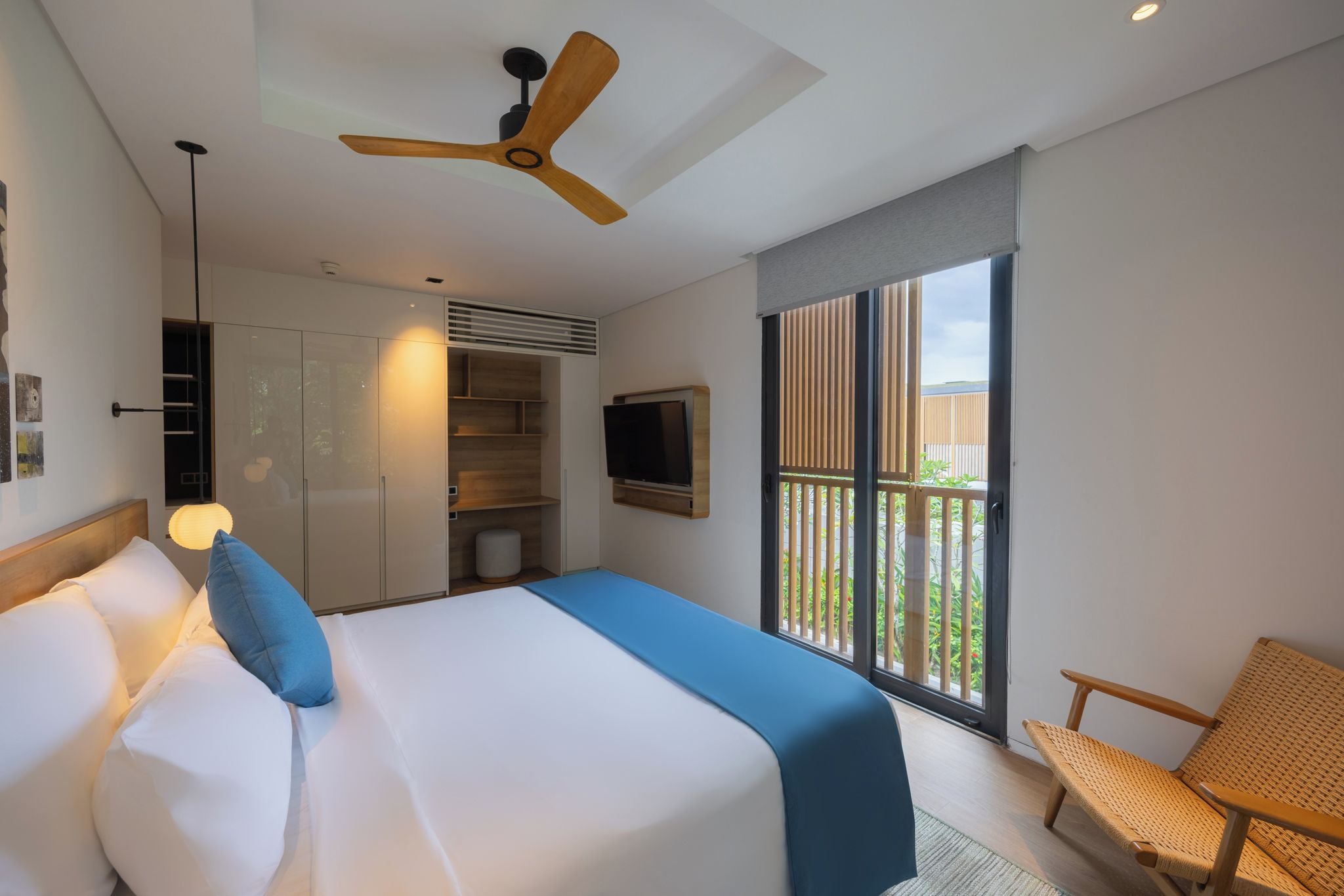 Quarto de hotel com cama, ventoinha no teto, televisão, varanda com vista e uma cadeira.