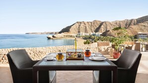 Uma mesa com vista para o mar e montanhas, duas cadeiras de vime, um tabuleiro com comida e bebidas, e um vaso com planta.