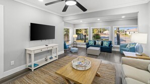 Uma sala de estar moderna com TV de tela plana, ventilador de teto e grandes janelas com vista para o exterior.