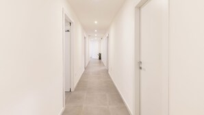 Um corredor comprido e estreito, com paredes brancas e chão de azulejos, que leva a uma porta no final.