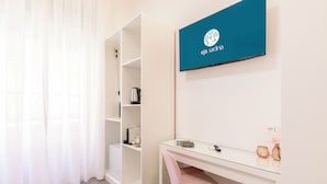 Quarto moderno com escrivaninha de vidro, cadeira rosa, estante branca e televisão na parede exibindo 'eja sardinia'.