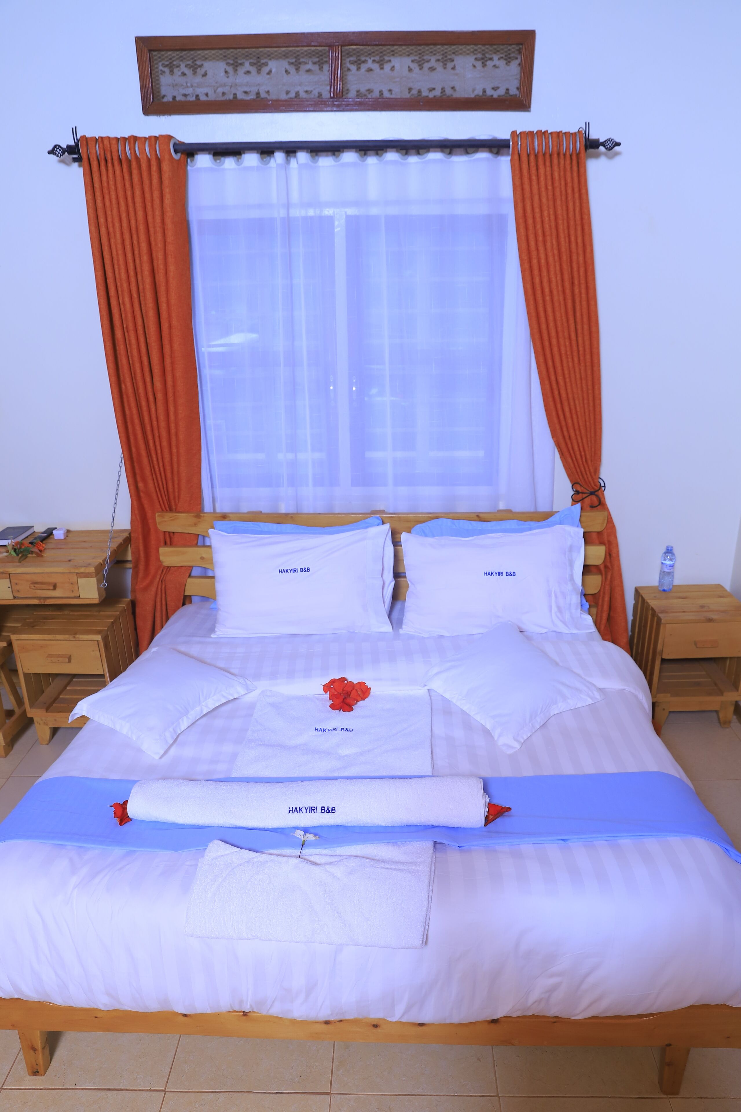 Cama bem arrumada com lençóis brancos e um cobre-leito azul, ladeada por duas mesinhas de cabeceira de madeira, com uma garrafa de água em uma delas.