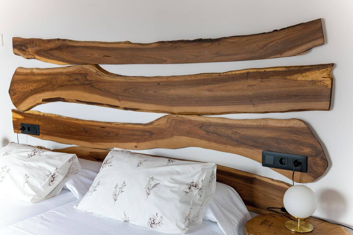 Uma cama com roupa de cama floral branca, uma cabeceira de madeira com tomadas elétricas embutidas e uma mesa de cabeceira com uma luminária de mesa branca em formato de globo.
