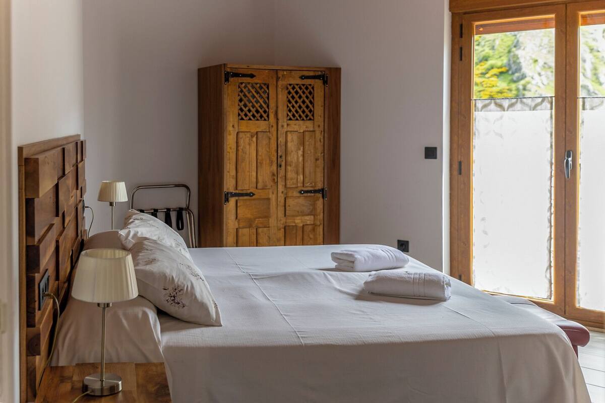 Quarto com guarda-roupa de madeira, cama com roupa de cama branca, luminária de cabeceira e vista para o verde através de uma porta de vidro.