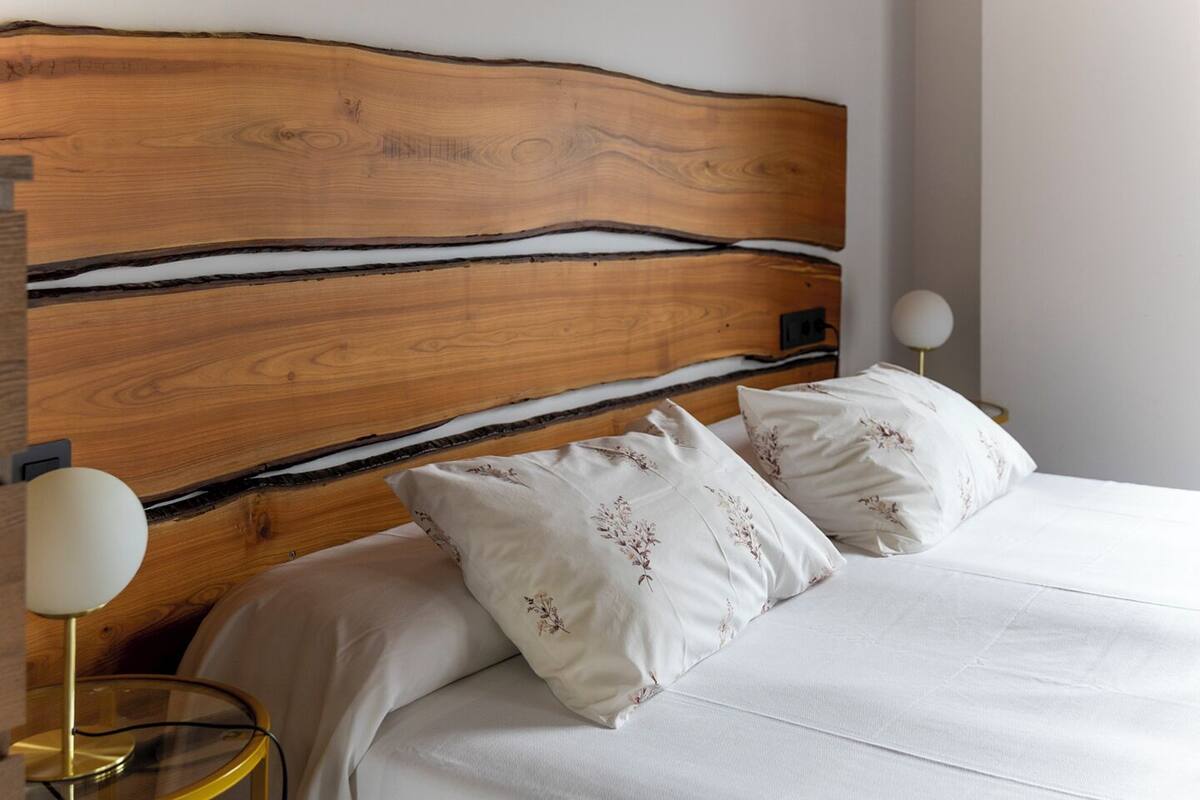 Uma cama com roupa de cama branca e almofadas florais, uma cabeceira de madeira com design exclusivo e uma mesa de cabeceira com abajur.