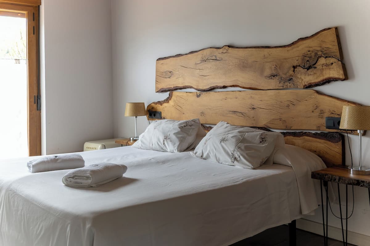 Cama bem arrumada com lençóis brancos e duas almofadas, cabeceira de madeira, abajur e uma pequena mesa com luminária.