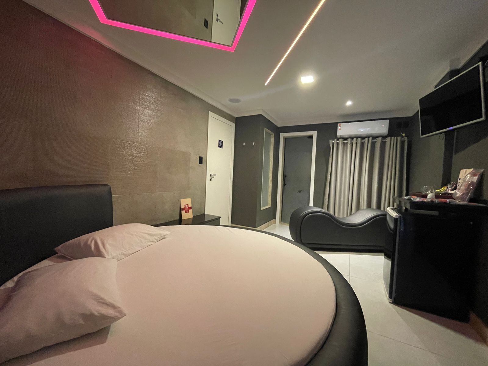 Quarto de hotel moderno com cama grande, escrivaninha, cadeira e televisão fixada na parede.