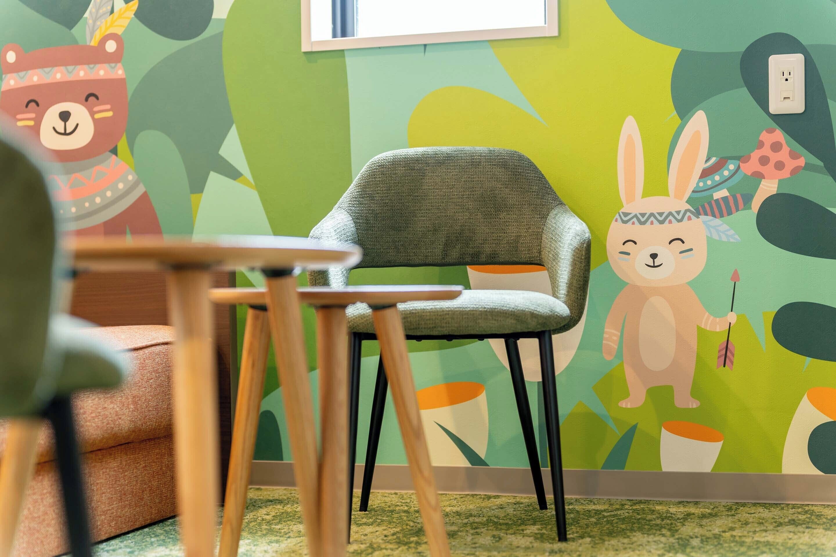 Quarto com parede verde com um mural de urso e coelho, uma cadeira cinza, uma mesa redonda pequena e piso de madeira.