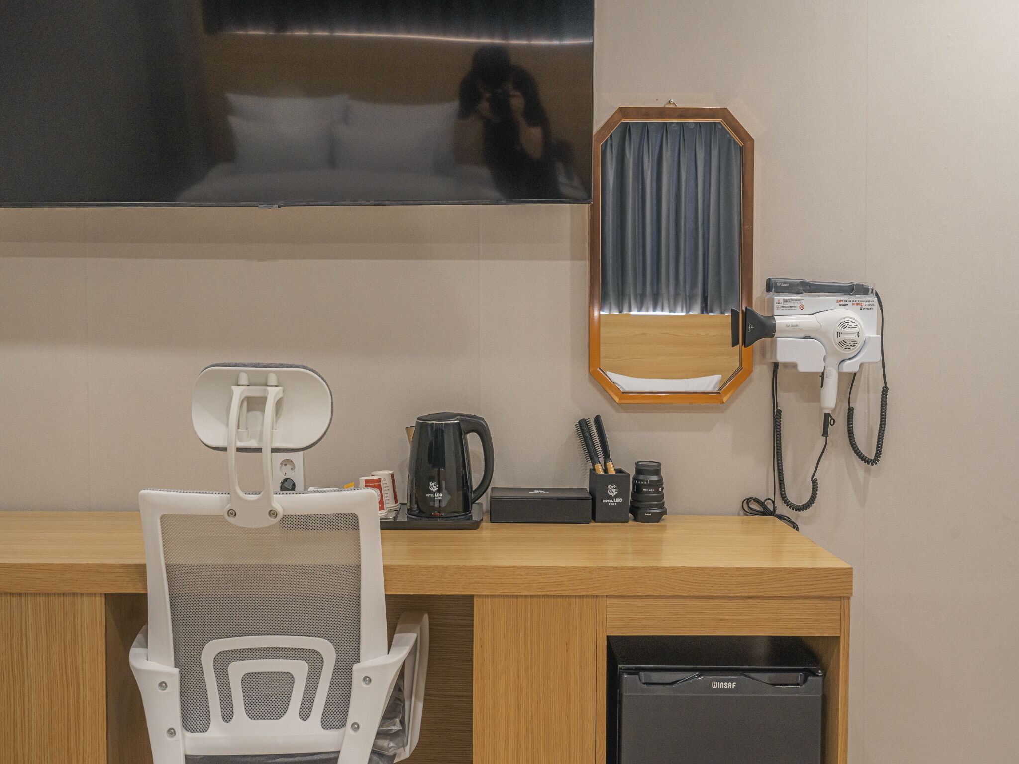 Quarto de hotel com escrivaninha de madeira, uma cadeira branca, televisão fixada na parede, espelho, secador de cabelo e um pequeno armário.