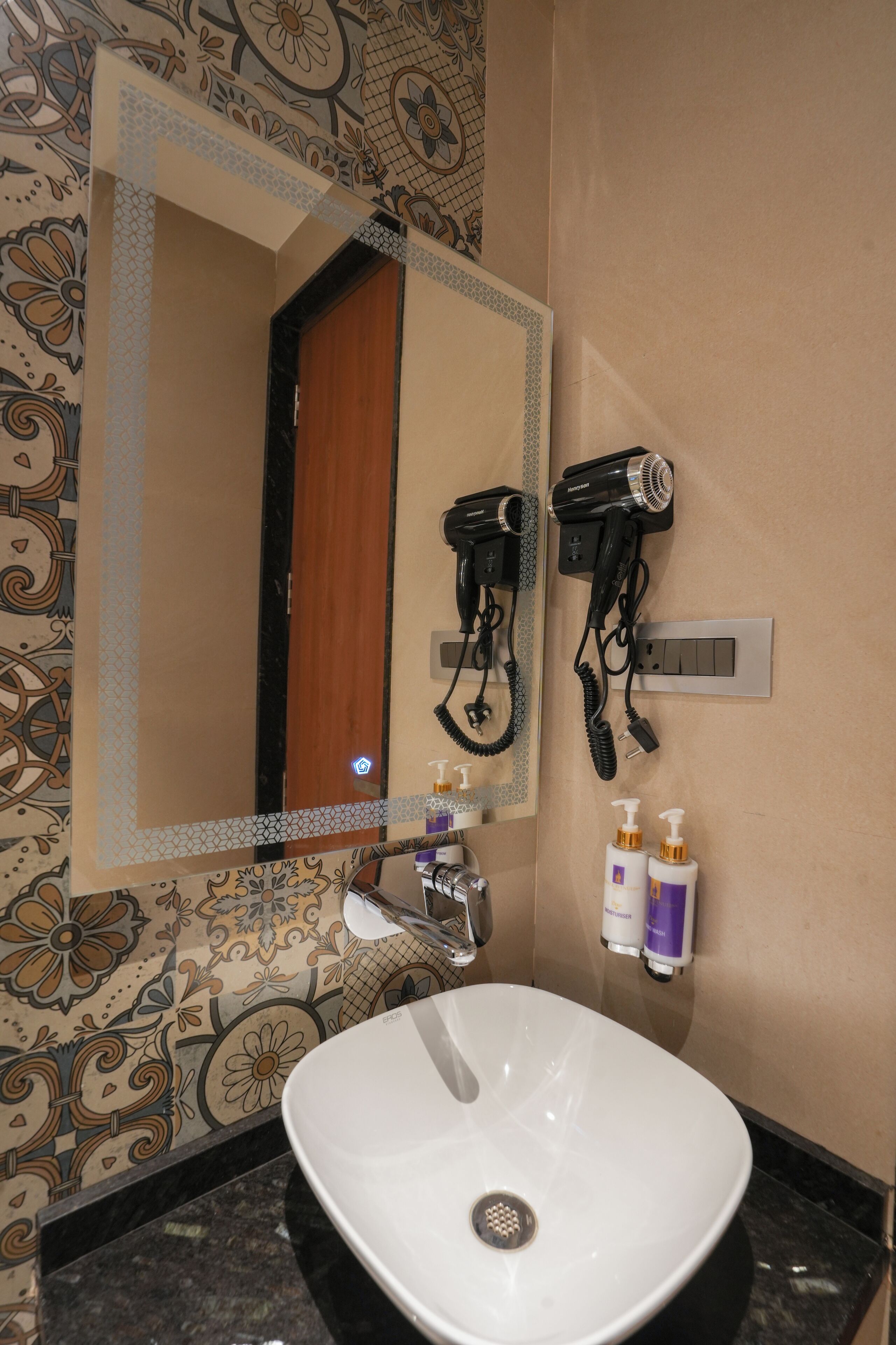 Casa de banho com lavatório branco, espelho com moldura decorativa, secador de cabelo e duas embalagens de gel desinfetante para as mãos.