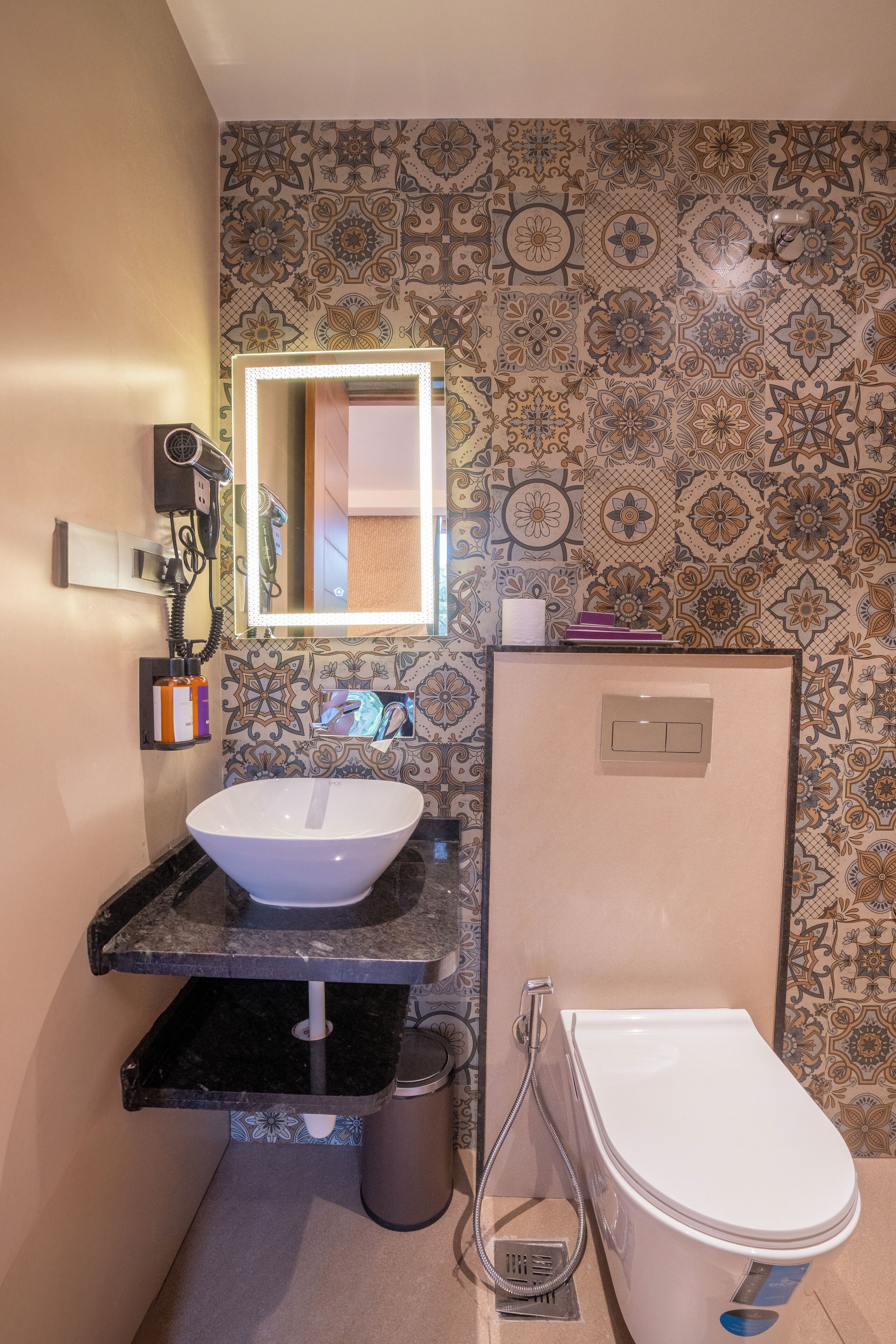 Um banheiro com pia branca, espelho com iluminação e vaso sanitário, além de azulejos estampados nas paredes.