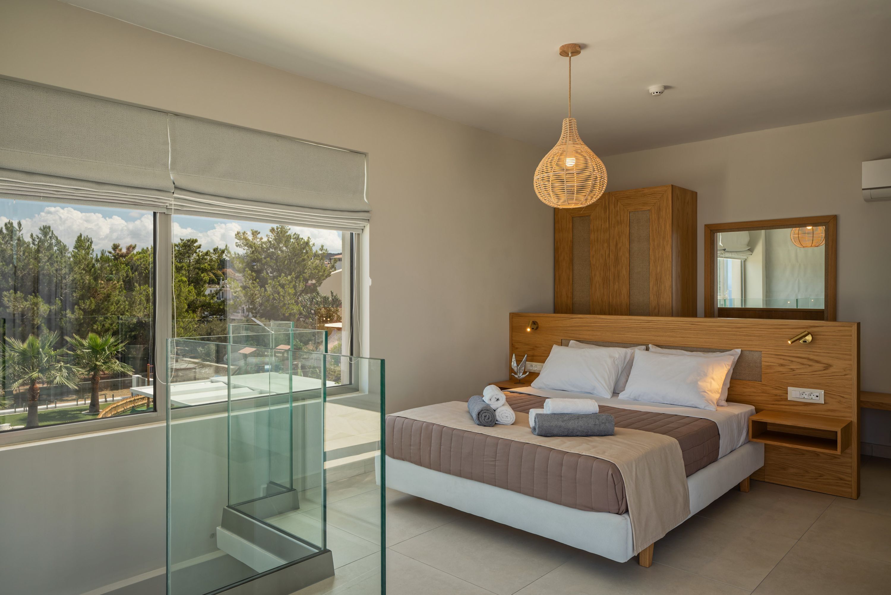 Quarto moderno com cama grande, cabeceira de madeira, varanda de vidro e luminária pendente.