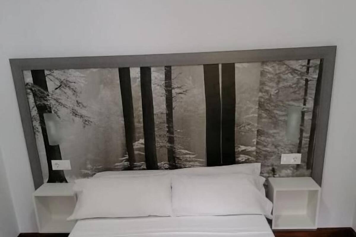 Cama bem arrumada com lençóis brancos e uma toalha enrolada, cabeceira com um mural de floresta e mesas de cabeceira brancas.