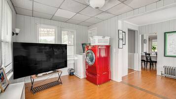 Sala de estar com televisão de tela plana, máquina de lavar vermelha, piso de madeira, paredes brancas e ventilador de teto.