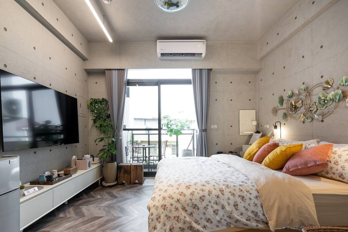 Quarto moderno com televisão de tela plana, cama com roupa de cama floral, planta em vaso e vista para varanda.