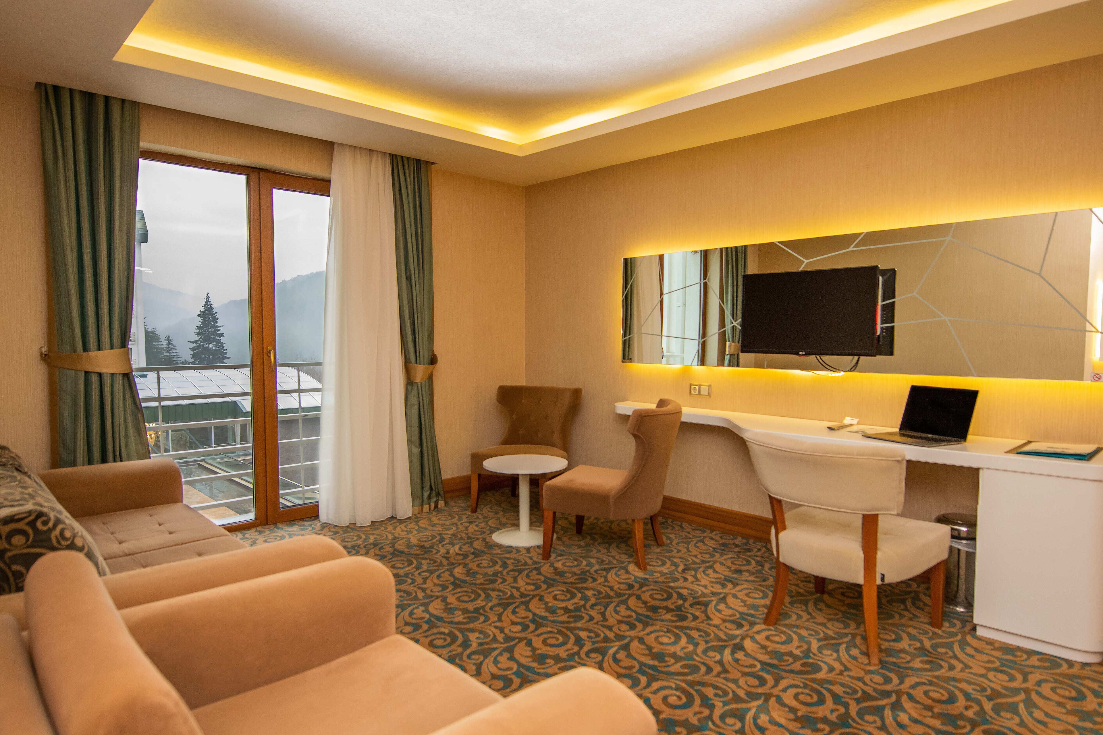 Quarto de hotel com sofá, duas cadeiras, escrivaninha com laptop, televisão de tela plana e uma janela grande com cortinas.