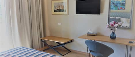 Quarto de hotel com cama, escrivaninha, cadeira, televisão, quadro e espelho.