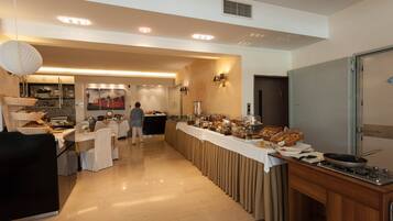 Buffet montado com diversas opções de comida, alguém servindo e uma área de cozinha com fogão.
