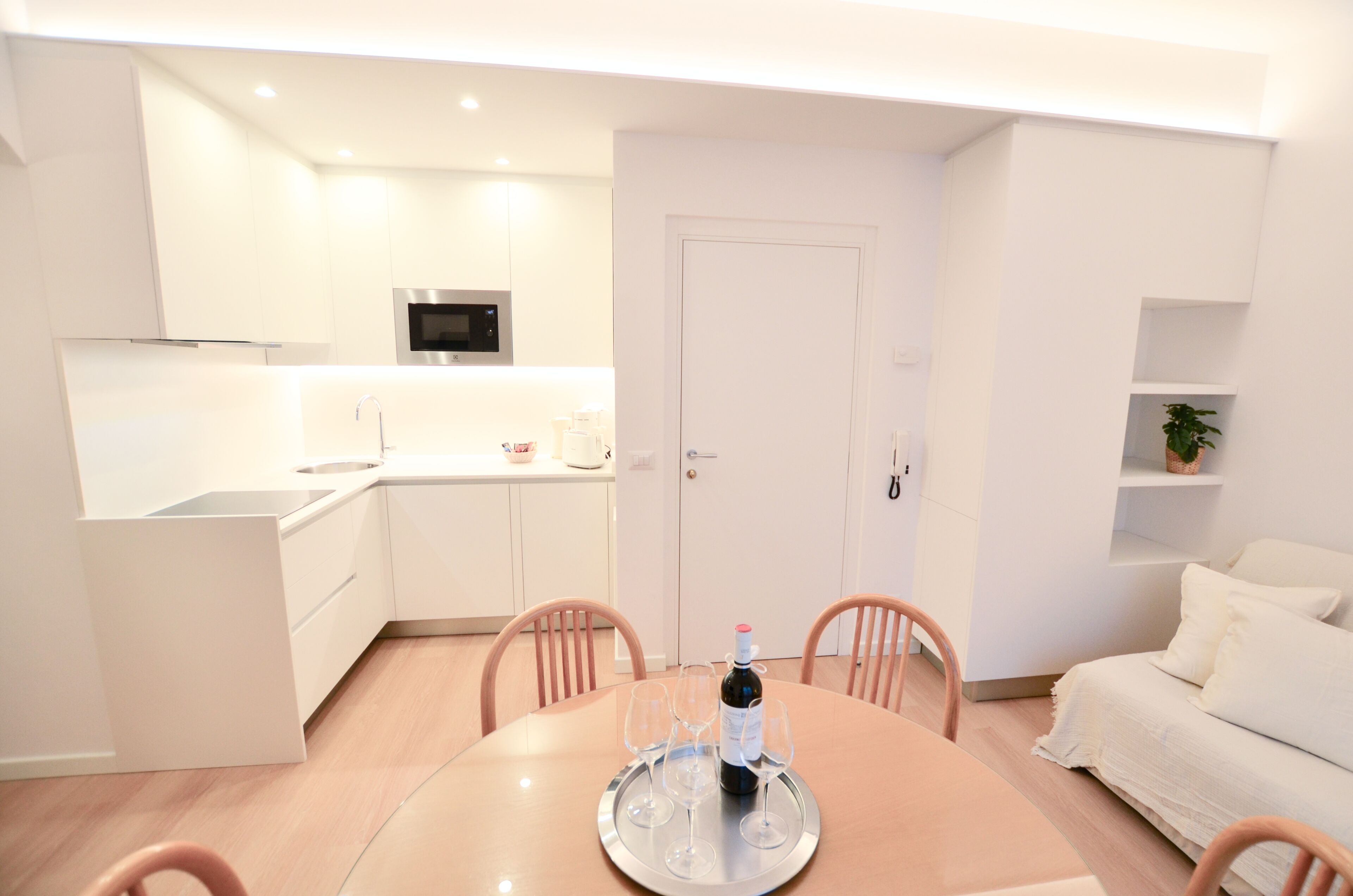Quarto moderno e bem iluminado com uma mesa de jantar posta para quatro pessoas, área de cozinha com armários brancos e um sofá branco.