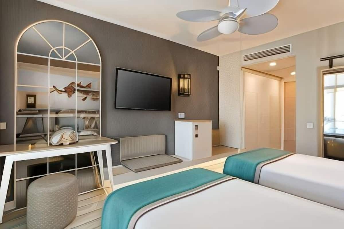 Quarto de hotel com cama, escrivaninha, televisão, ventoinha no teto e guarda-roupa.