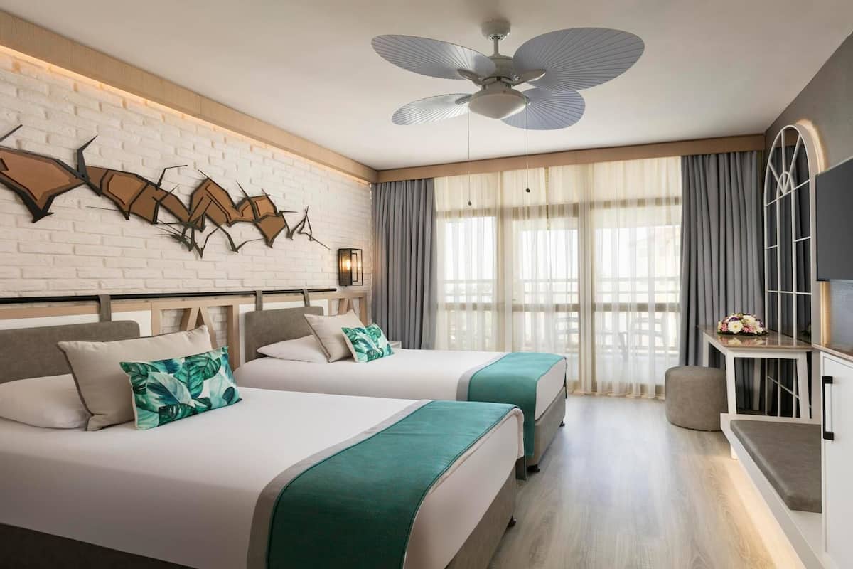 Quarto de hotel com duas camas, ventilador de teto, televisão e parede de tijolos com detalhes decorativos.