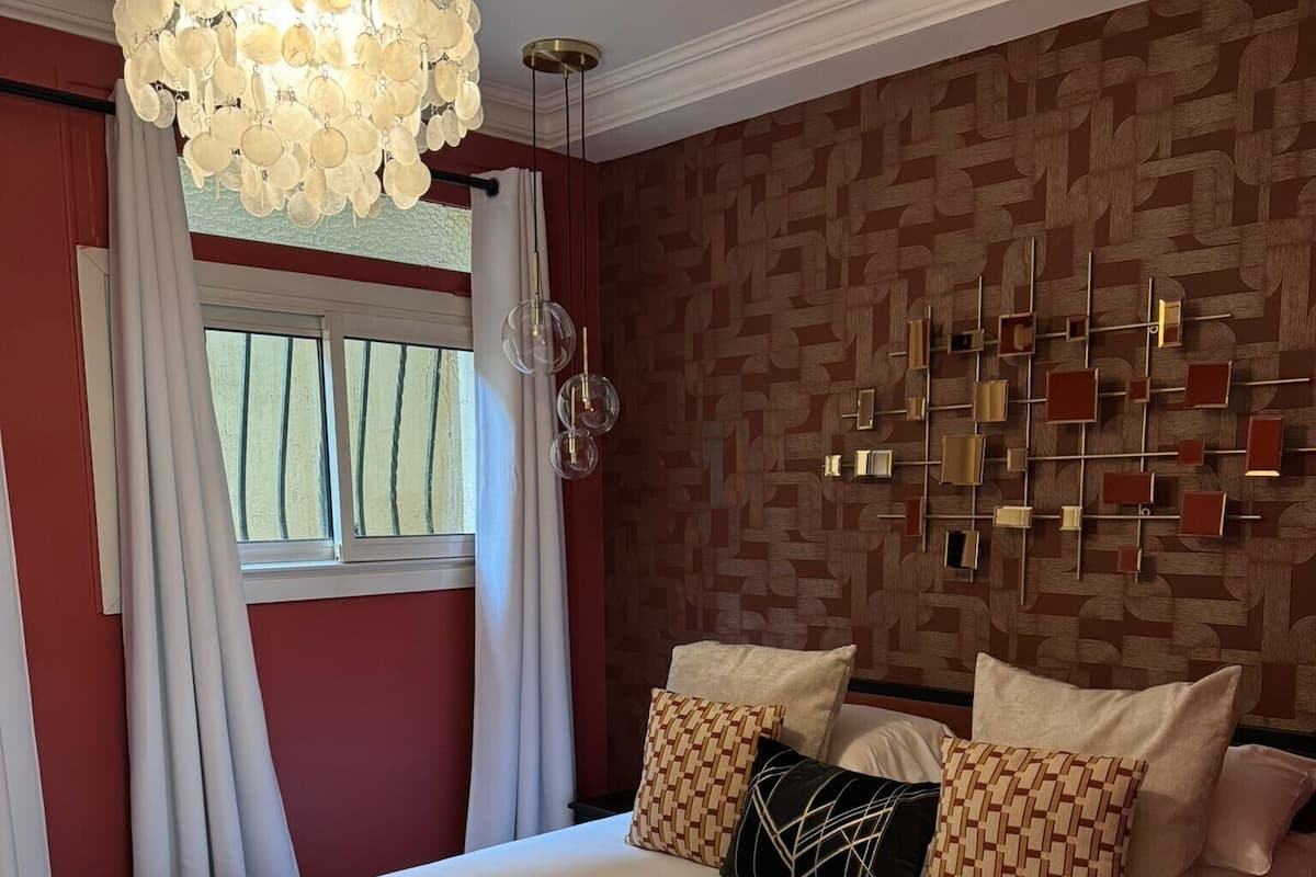 Quarto de hotel com uma cama grande, um painel decorativo na parede, um candelabro e uma janela com cortinas.