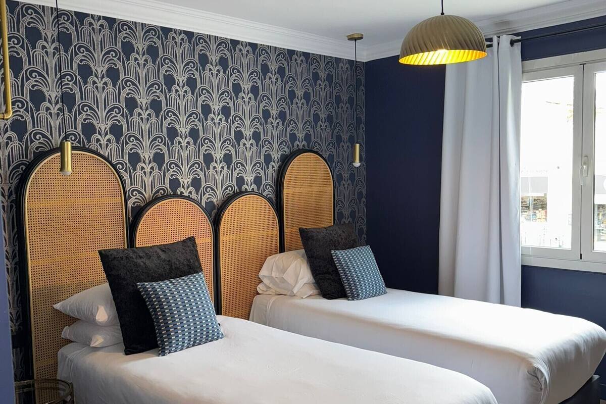 Quarto de hotel com duas camas, papel de parede estampado, teto com uma medalhão decorativa e janela com cortinas.