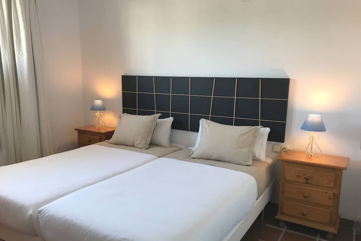 Uma cama de casal com cabeceira, duas mesas de cabeceira com abajures e uma janela com cortinas.