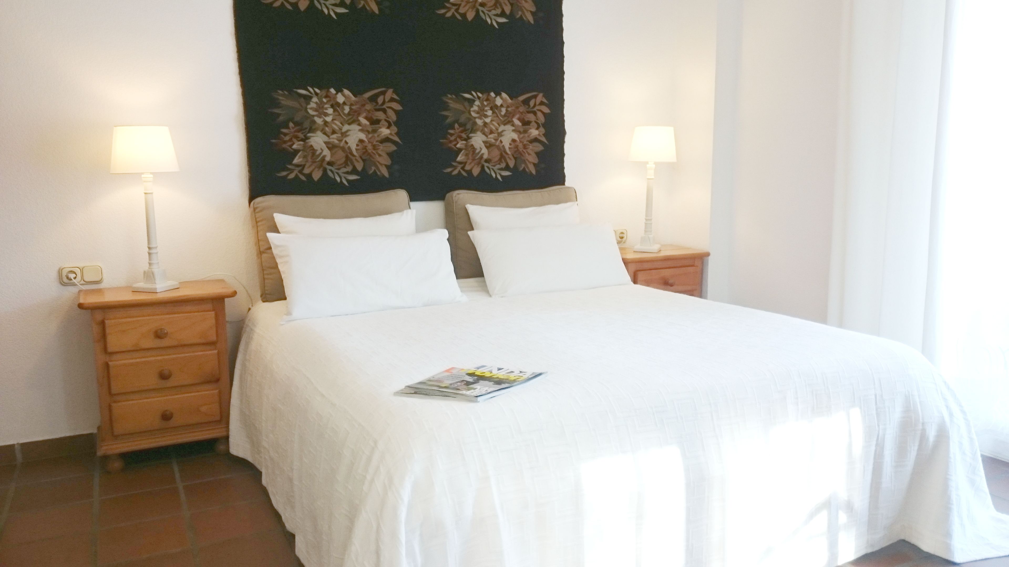 Cama bem arrumada com lençóis brancos, duas luminárias de cabeceira e uma revista sobre a cama.