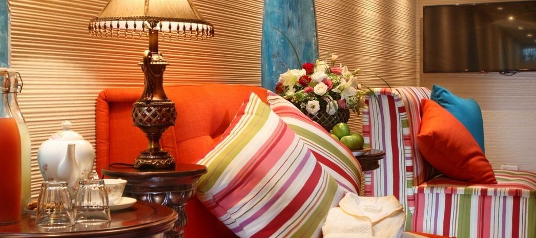 Uma sala de estar aconchegante com almofadas listradas, uma mesa com abajur, um vaso de flores e uma televisão na parede.