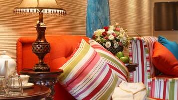 Uma sala de estar aconchegante com almofadas listradas, uma mesa com abajur, um vaso de flores e uma televisão na parede.