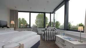 Quarto de hotel moderno com uma cama grande, área de estar com cadeiras listradas, escrivaninha com abajur e janelas amplas com vista para árvores.