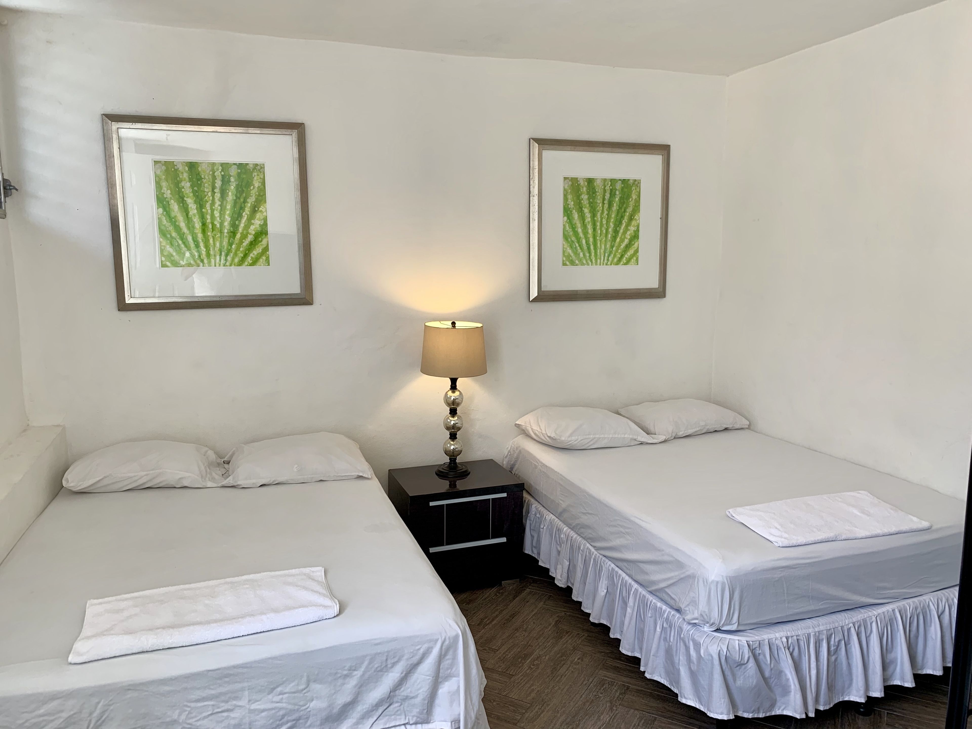 Quarto de hotel com duas camas, uma mesa de cabeceira com abajur e duas molduras na parede.