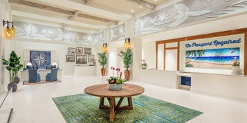 Um hall de entrada com balcão de recepção, quadros decorativos, uma mesa redonda de madeira com plantas e uma parede com o aviso 'No Passport Required'.