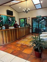 Uma área de recepção com balcão de madeira, um candelabro, uma televisão montada acima do balcão e uma grande janela com vista para vegetação.