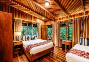 Quarto com painéis de madeira, duas camas, uma mesa de cabeceira com abajur e uma janela com cortinas.