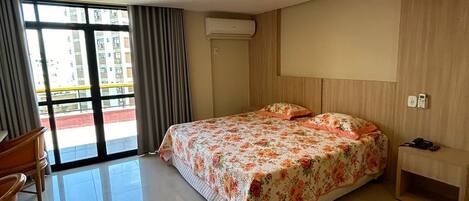 Quarto de hotel com cama, escrivaninha, cadeira, janela e ar condicionado.