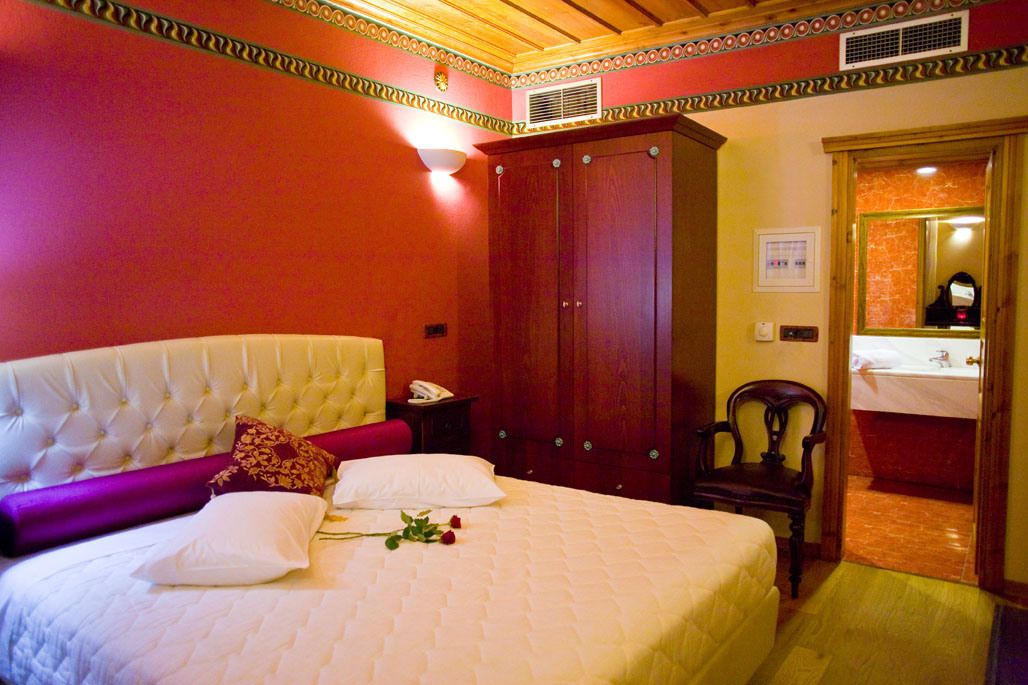 Quarto de hotel com uma cama grande, um guarda-roupa de madeira, uma cadeira e um banheiro visível através de uma porta aberta.