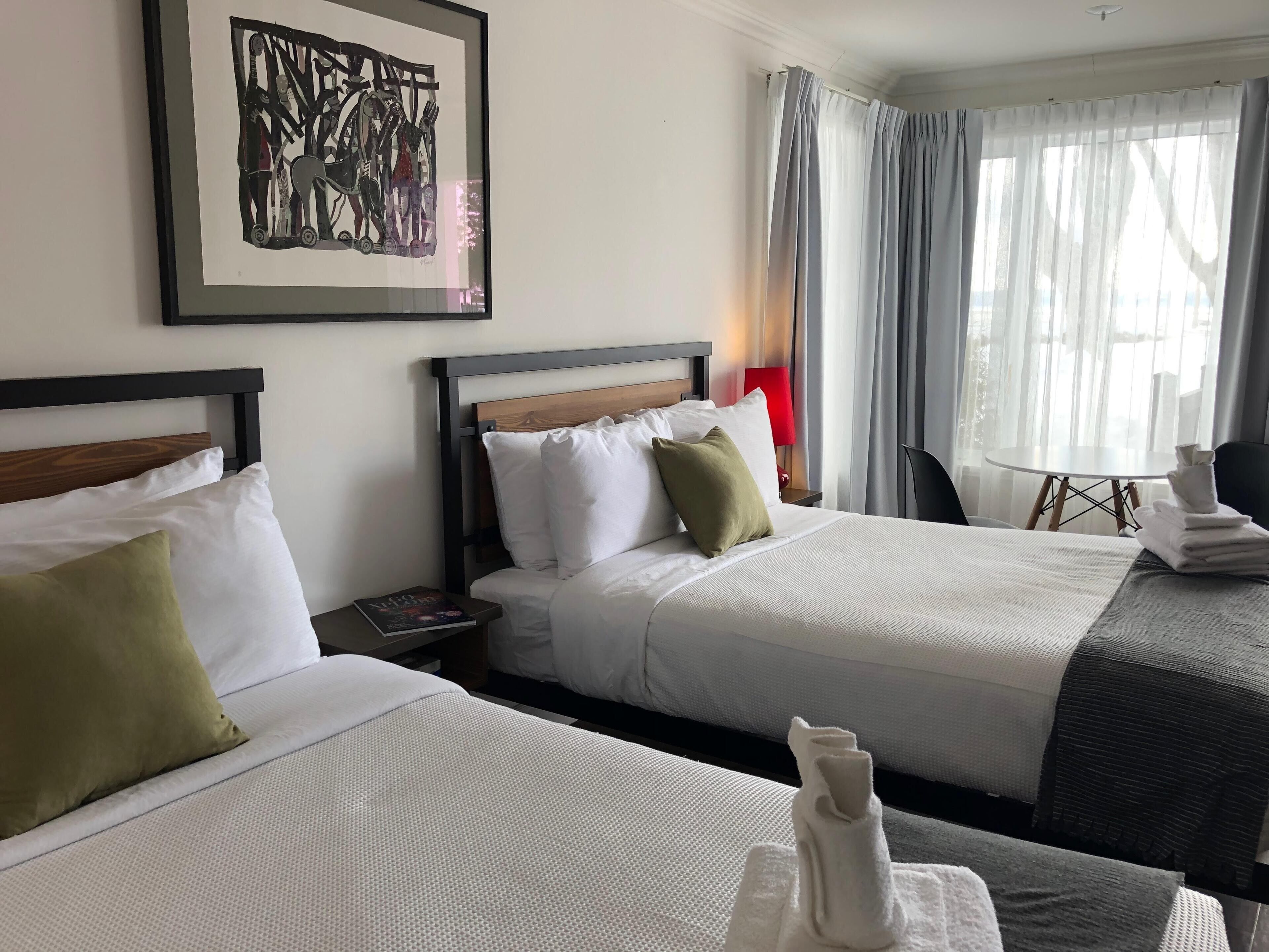 Quarto de hotel com duas camas, uma obra de arte emoldurada na parede, uma mesinha de cabeceira com abajur e vista para o exterior através de cortinas translúcidas.
