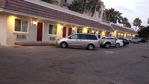 Um motel com uma fileira de quartos, cada um com porta e janelas, e um estacionamento com vários carros estacionados em frente.