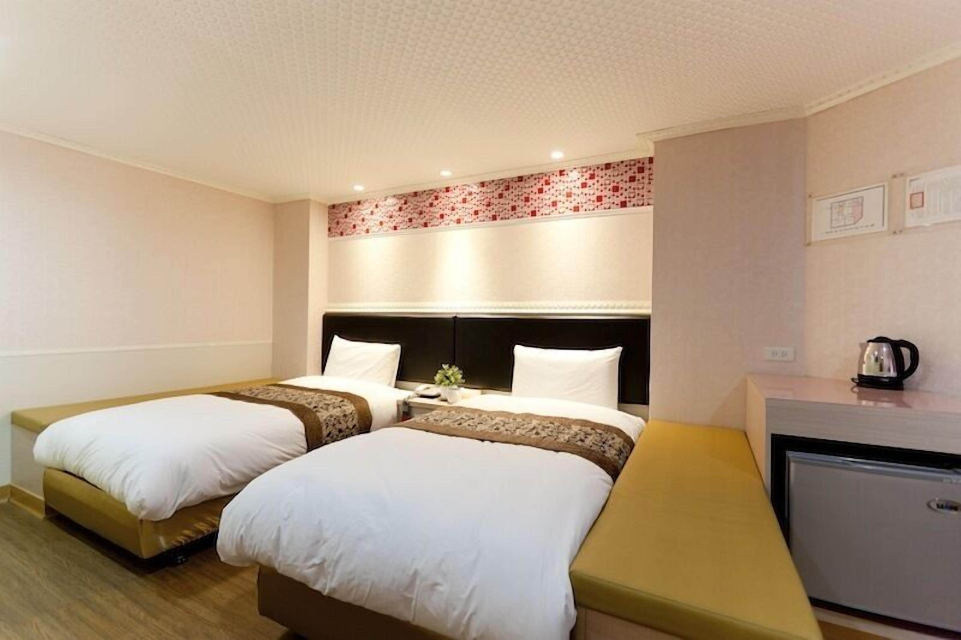 Quarto de hotel com duas camas de solteiro, uma pequena mesa com chaleira e um refrigerador.