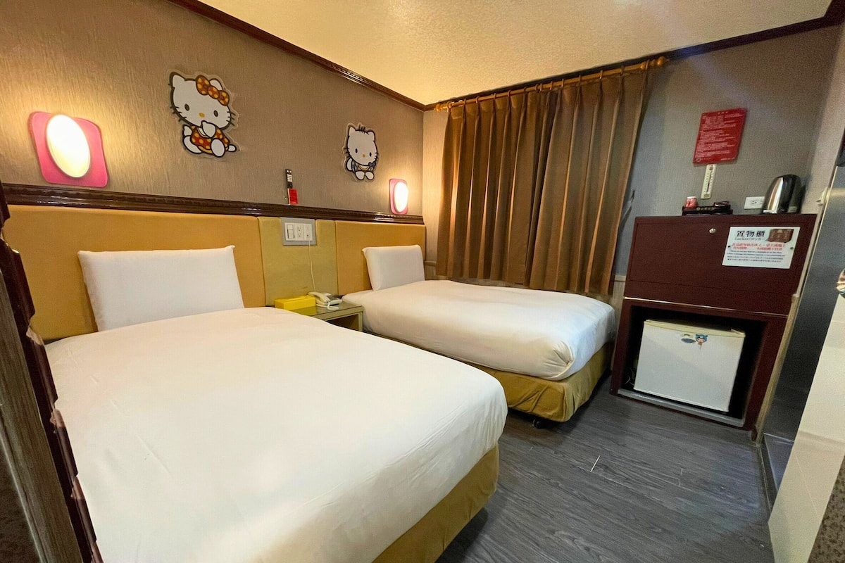 Quarto de hotel com duas camas, uma pequena mesa, uma mesinha de cabeceira e uma decoração de Hello Kitty na parede.