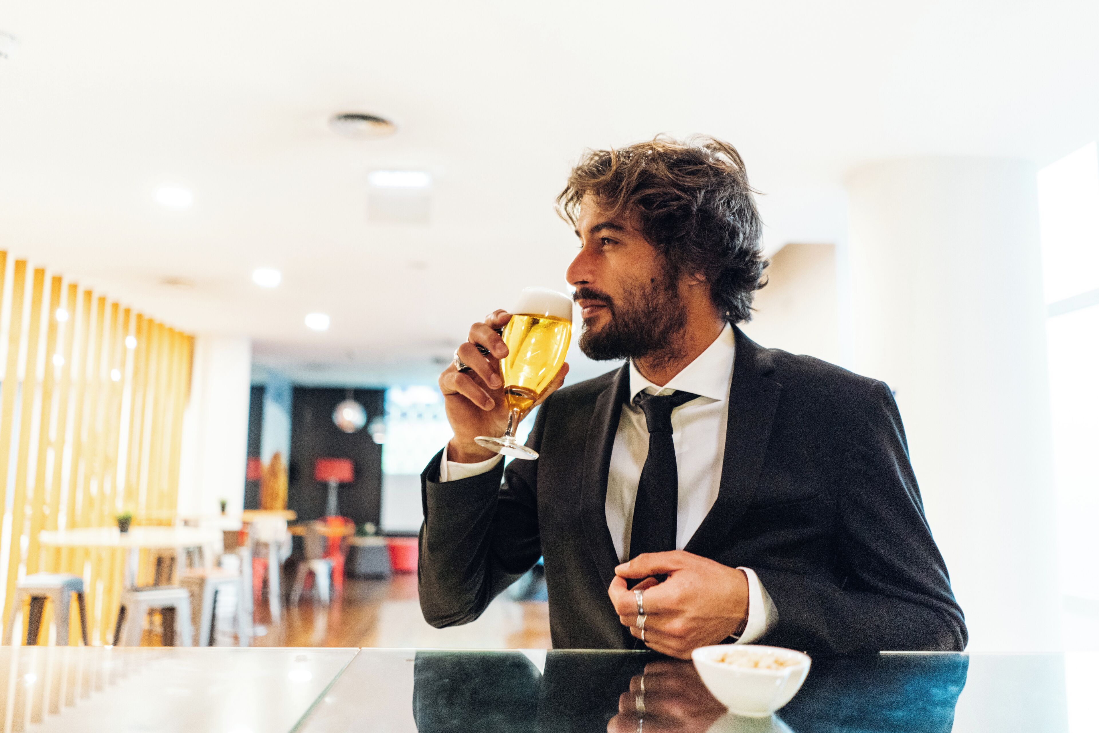 Um homem de terno segura um copo de cerveja num bar com decoração moderna e detalhes em madeira.