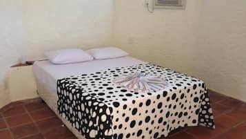 Quarto com cama, paredes brancas, colcha de bolinhas e piso ladrilhado.
