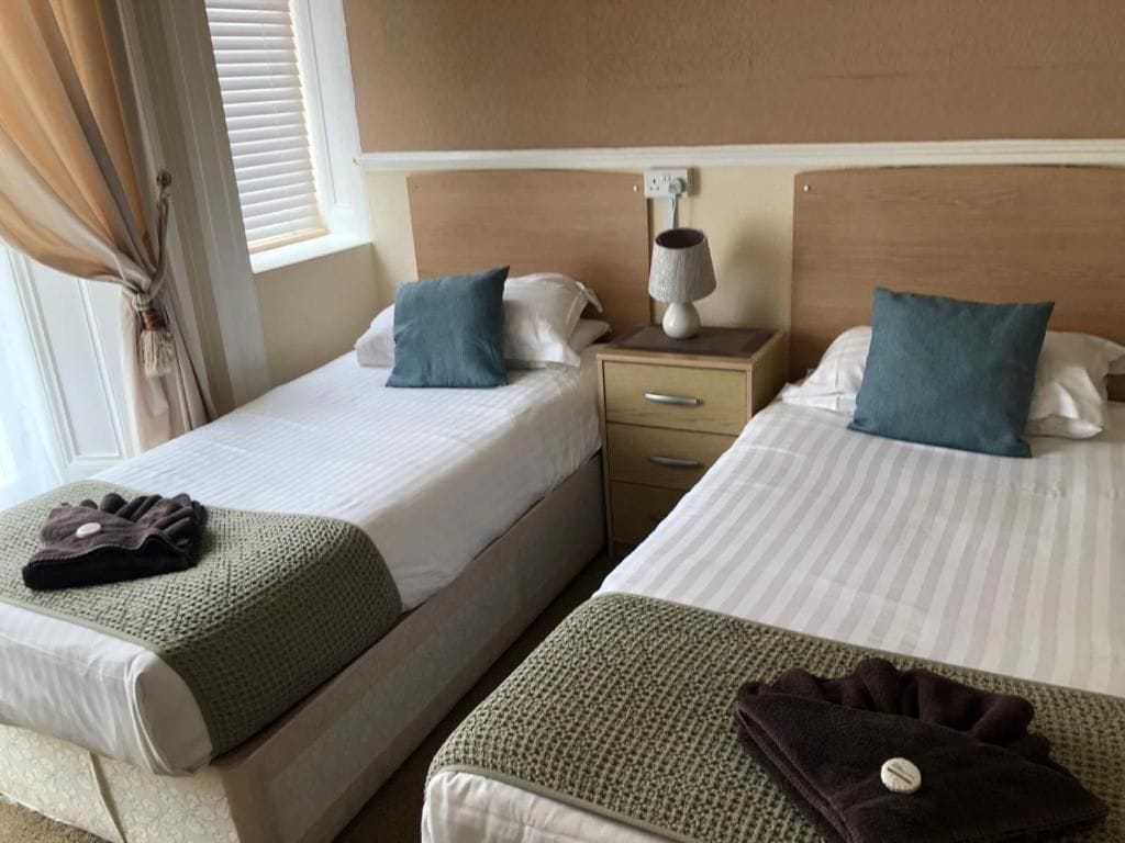 Quarto de hotel com duas camas, mesa de cabeceira com abajur e janela com cortinas.